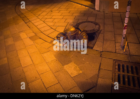 bronze statue of a man playfully peeking out from under a manhole cover ...