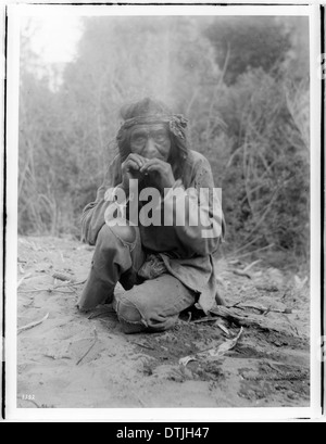 An elderly Havasupai Indian man crouching and rolling a cigarette ...
