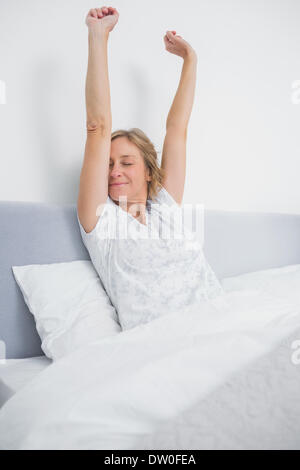 Well rested blonde woman stretching after waking up in bed in bedroom ...