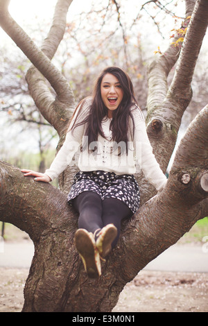 Brunette woman sitting on a tree in a green grass field, looking at her ...