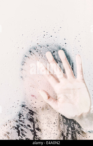 Asian woman behind window pressing hand against pane with water drops ...
