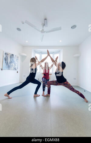 three women partake in a group yoga fitness class, in an empty room ...
