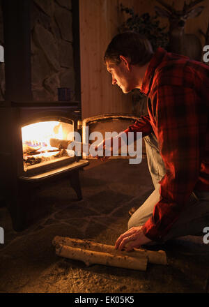 mid-adult man adding logs to a wood burning stove fire in rustic ...