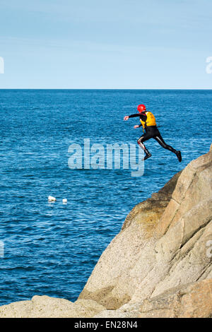 Man in wetsuit, helmet and lifejacket jumping off rocks into sea ...