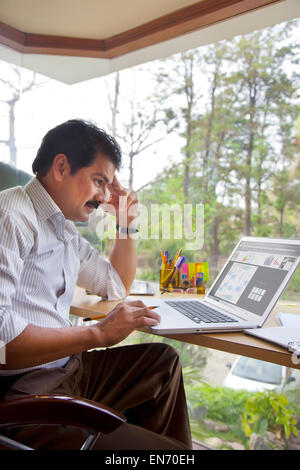 Thinking while holding pencil, man working on laptop at home office ...