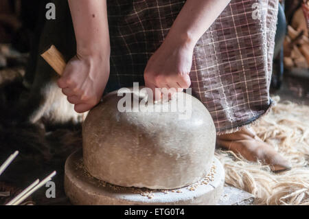 A young woman dressed in iron-age clothes grinds wheat using a heavy ...