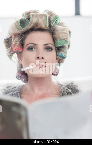 Portrait of woman with hair curlers and magazine smoking a cigarette ...