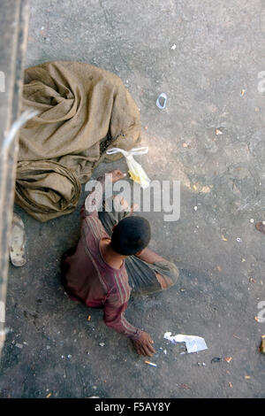 A scavenger man living in poverty sits on a sidewalk while he sniffs ...