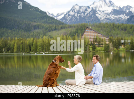 A senior couple with their dog sitting on the wooden dock over the lake ...