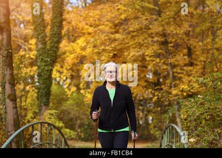 Senior female nordic walker crossing over footbridge in autumn park ...