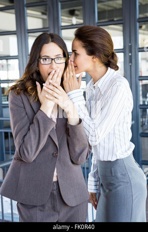 An office worker whispering in the ear of a colleague, with a shocked ...