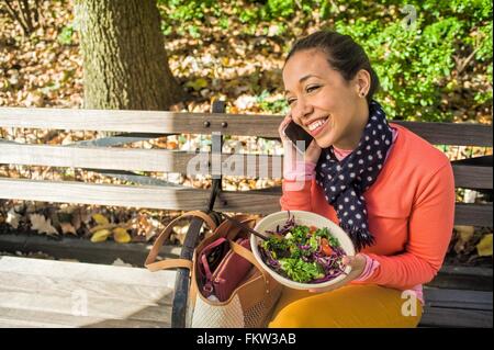 Woman sitting on a bench, eating from a plate of food balanced on her ...