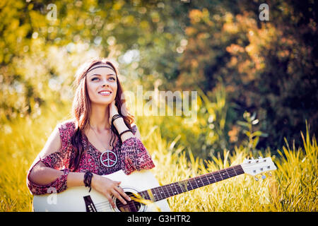 outdoor portrait of beautiful hippie girl sitting on green grass near birch with guitar. lake ...