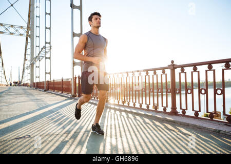 Attractive fit man running fast along big modern bridge at sunset light ...