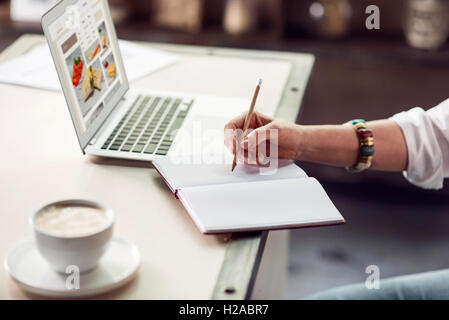 Hand of female chef writing recipe in diary by vegetables and cutlery ...