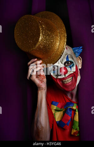 a scary evil clown peering out from a purple stage curtain, with his ...