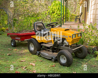 Yellow ride-on mower towing small red trailer full of soil on lawn ...