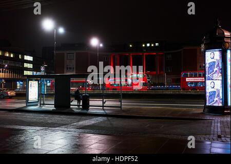 Woman sitting at a bus stop at night, looking at her phone with city ...