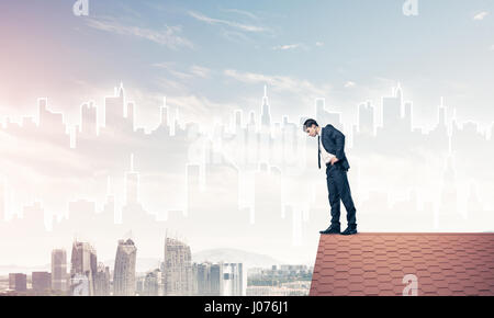 Businessman looking down from roof and modern cityscape at background ...