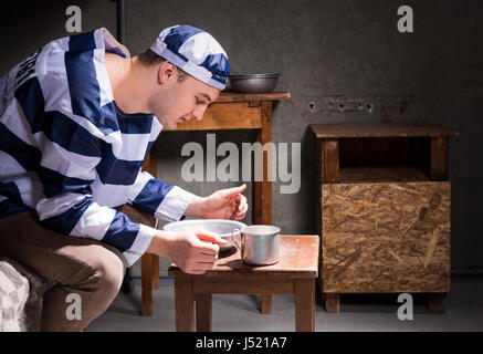 Young man wearing prison uniform sitting on a bed near bedside table ...