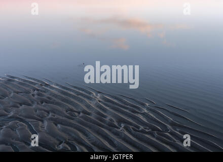 Reflections of drifting clouds bounce off smooth water on rippled beach ...