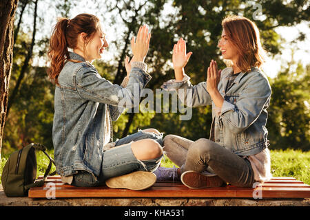 Two pretty girls clap their hands while sitting on wooden bench in park ...