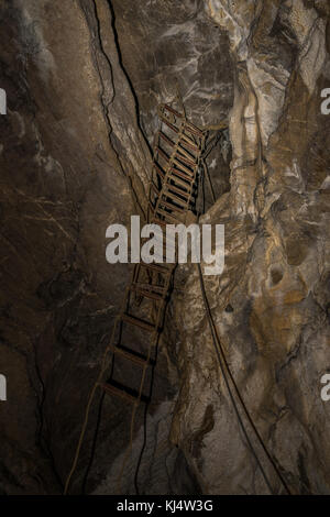 Wood ladder going up from a rocky stream bed through a narrow hole in a ...