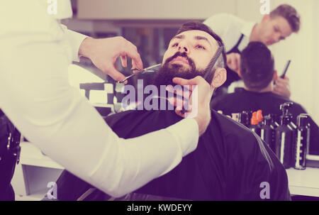 Cheerful man hairdresser forming beard of client into new shape at hair ...