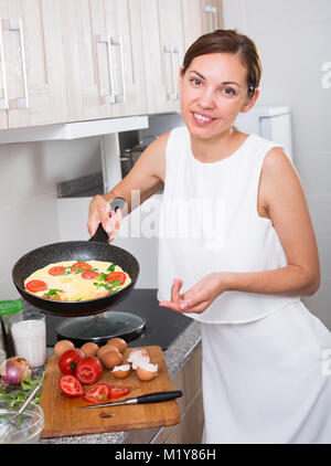 glad young woman holding frying pan with omelet at kitchen unit indoors ...