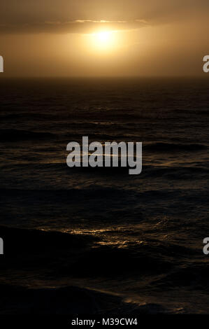 The sun sinks behind ocean waves as scattered rocks and warm light ...