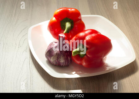 Two red bell peppers and a head of blue garlic lie on a white plate ...