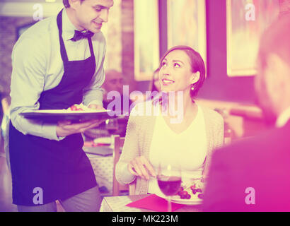 Diligent friendly smiling waiter with dishes serving man and woman ...