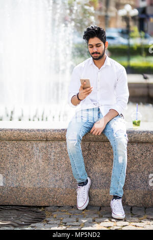 Young handsome indian man speak on phone sitting near fountain in the ...