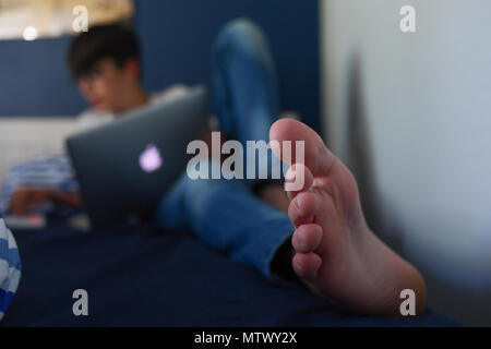 A teenage boy (AGE 14) lays on his bed with a mac book air laptop doing ...