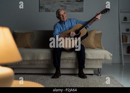 Senior happy man plays acoustic guitar while sitting on sofa in room ...