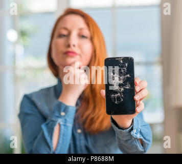 Redhead woman holding broken smartphone with a confident expression on ...