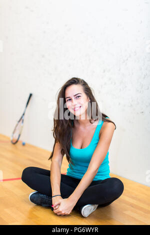 Cute young woman with a racket leaning against a wall in a squash court ...