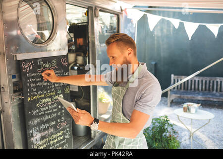 Male waitress writing menu on menu board while using digital tablet ...