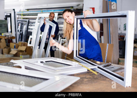 Smiling cheerful workwoman with plastic window frame in workshop giving ...