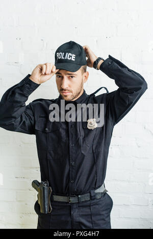 handsome young policeman putting on cap in front of white brick wall ...