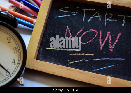 Start now phrase colorful handwritten on chalkboard, alarm clock with ...