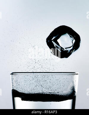 An ice cube dropping into a glass of water creating a splash. The glass has a blue tint at the ...