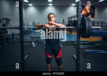 Muscular athlete in sportswear poses at the stand with barbell in gym ...