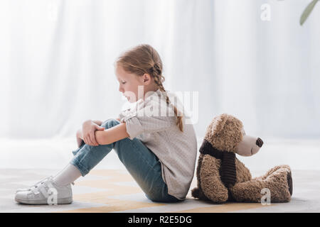 Side view of lonely sad little boy sitting on bed with hugging knee and ...