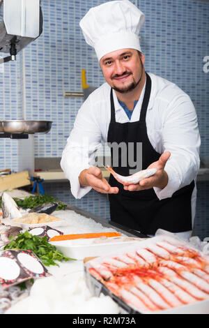 Handsome seller in white cap shows hands fish on counter in supermarket ...