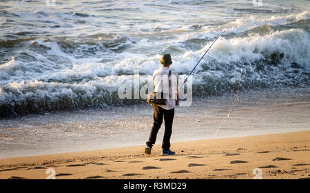 Fisherman stands with a fishing pole near the incoming ocean waves at ...
