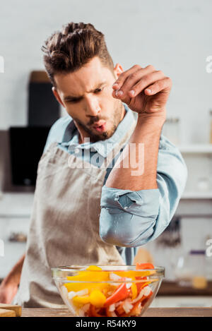 Cooking process. Man adding salt into bowl of salad at countertop in ...