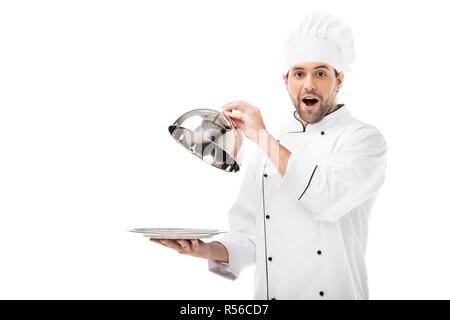 surprised young chef taking of serving dome from plate isolated on ...