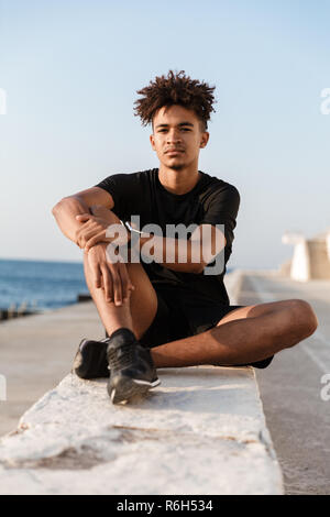 Image of concentrated young guy sportsman sitting outdoors on the beach ...