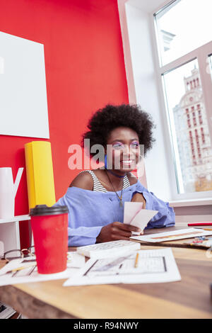 Dark-haired curly designer sitting near bright red wall and working ...